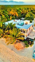 Uma propriedade à beira-mar com um edifício de dois andares, um alpendre com mobiliário de exterior e uma área de praia arenosa.