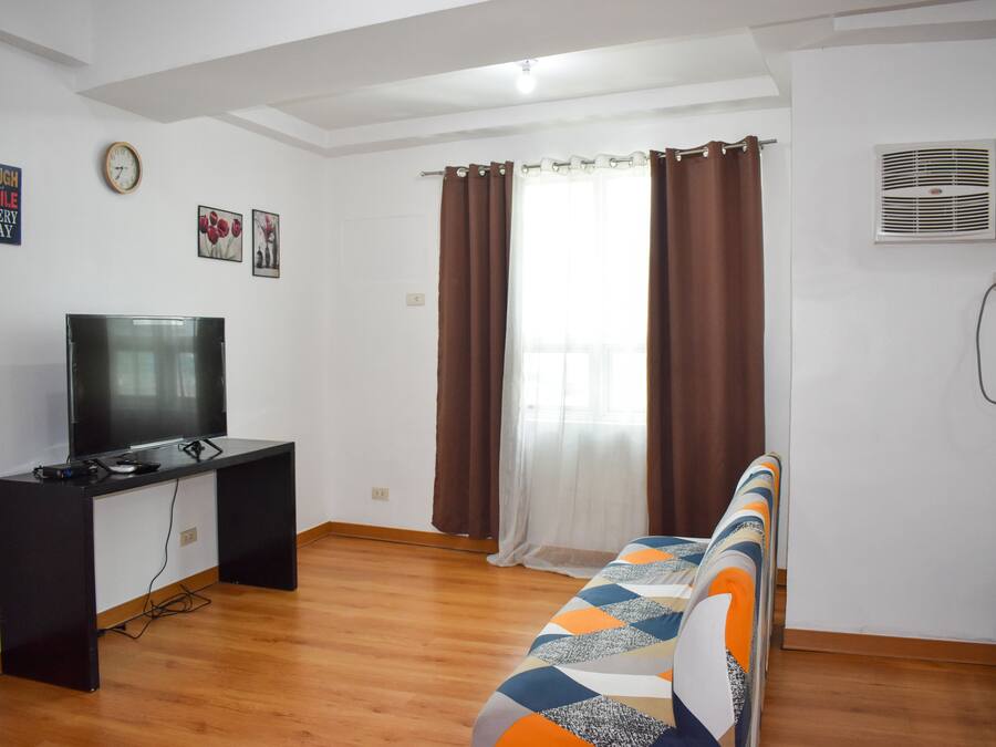 Quarto com televisão sobre uma escrivaninha, cama com manta colorida, janela com cortinas e ar condicionado fixado na parede.