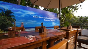 Restaurante com mesas e cadeiras de madeira, um cardápio e um painel ao fundo com uma cena de praia e texto.