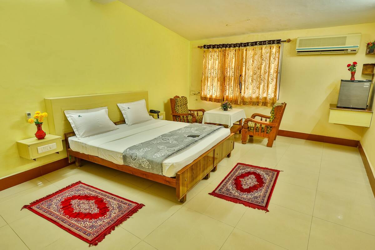Quarto de hotel com cama, mesa pequena, cadeira, mesa de cabeceira e janela com cortinas.