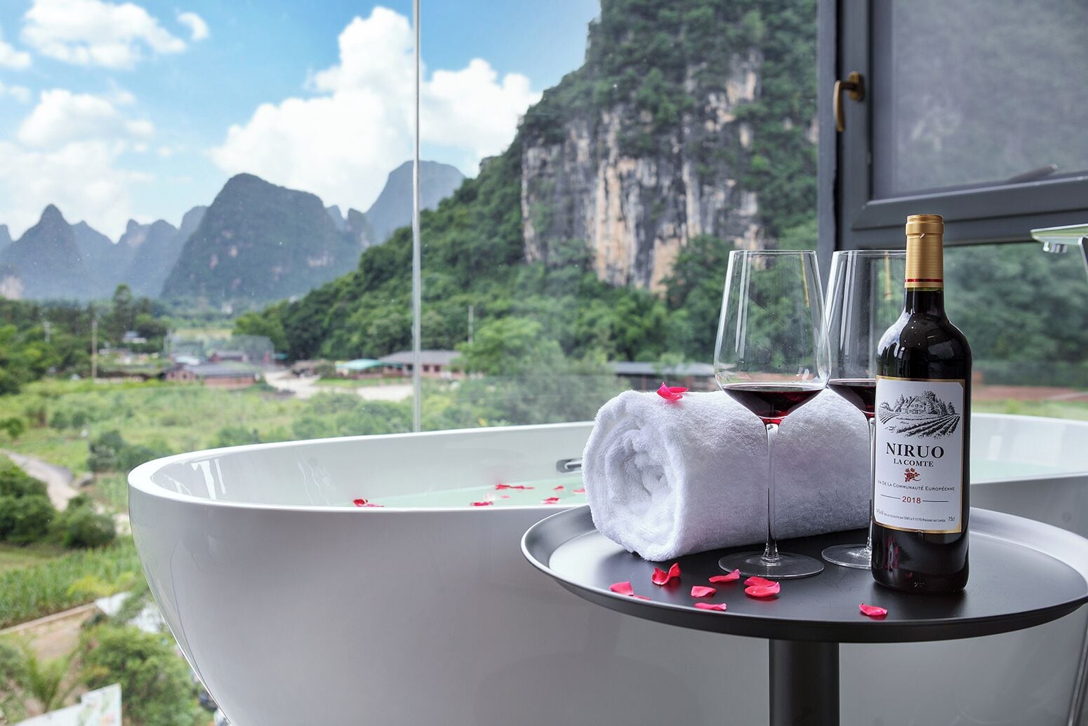 Uma banheira com uma garrafa de vinho e copos, rodeada por pétalas de rosa, com vista para montanhas.