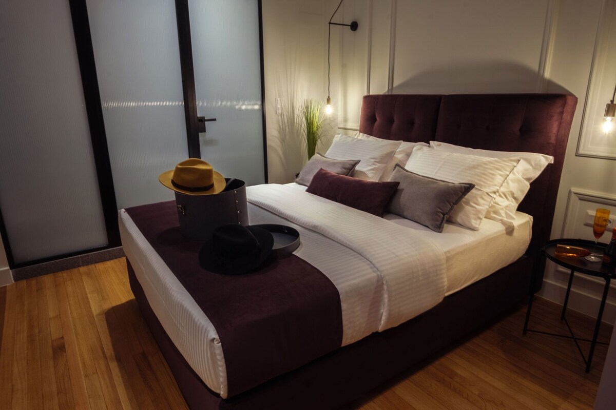 Uma cama com cabeceira escura, roupa de cama branca e alguns travesseiros. Há um chapéu e um pequeno recipiente na cama.