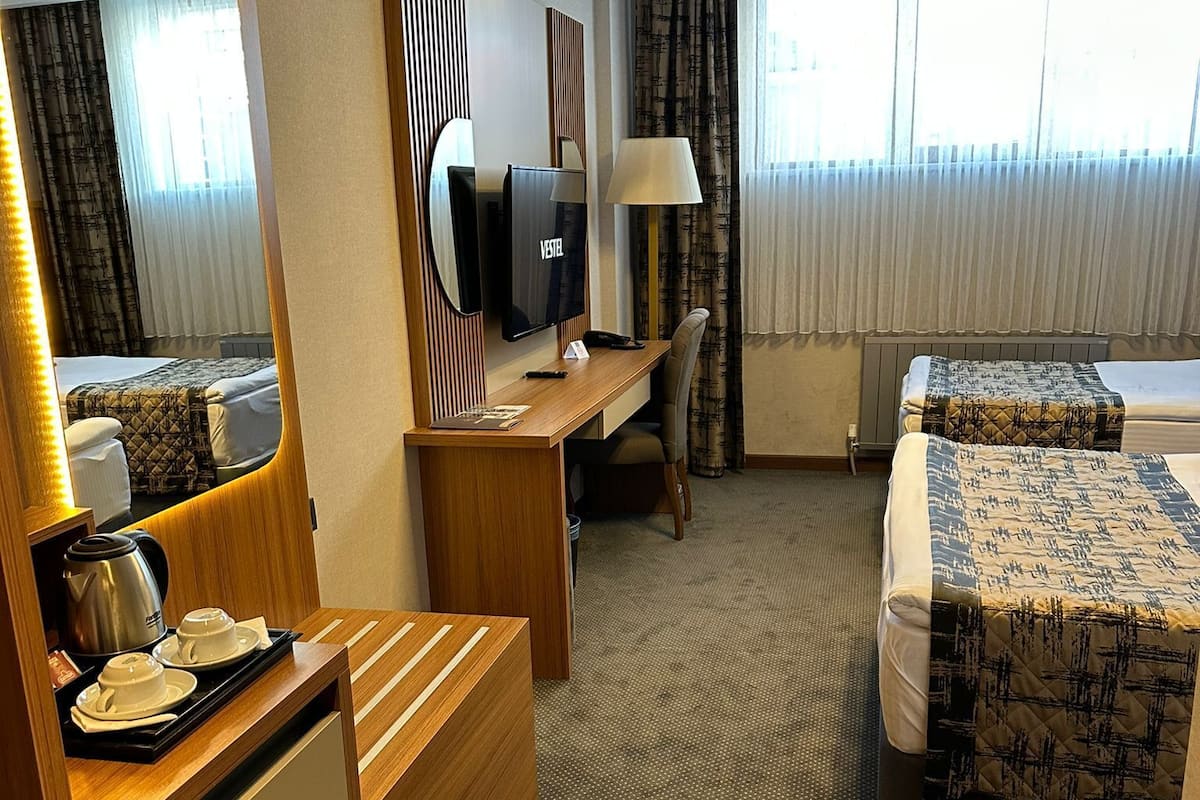 Quarto de hotel com uma cama, escrivaninha com computador, televisão, espelho e janela com cortinas.