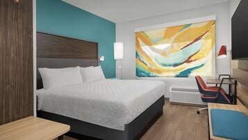 Quarto de hotel moderno com uma cama grande, uma escrivaninha, uma cadeira e um quadro abstrato colorido na parede.