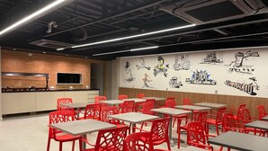 Restaurante com cadeiras e mesas vermelhas, área de cozinha e parede com diversos desenhos.