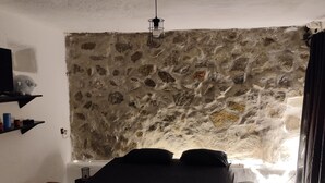 Quarto com parede de pedra, uma cama, uma mesinha e uma televisão em uma estante.