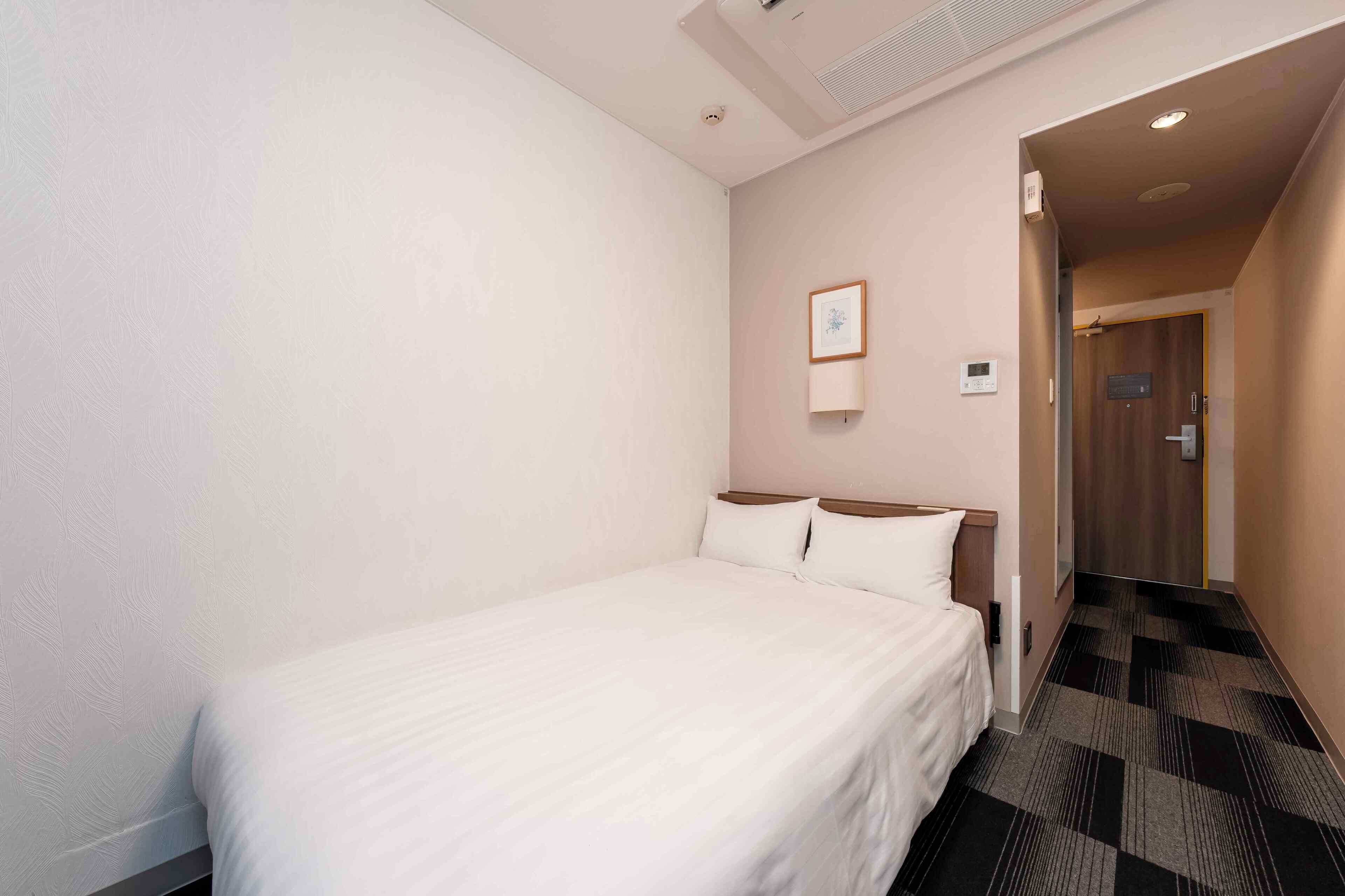 Quarto de hotel com uma cama de solteiro, mesa de cabeceira, luminária fixada na parede e porta que dá para outro quarto.