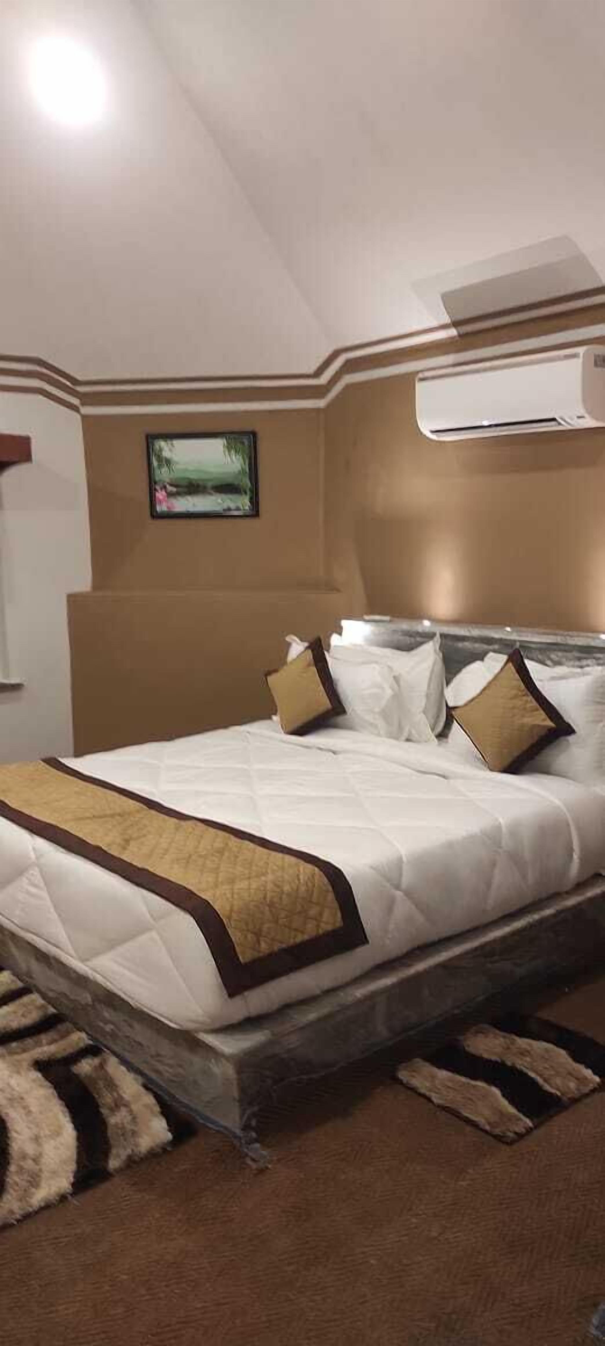 Cama bem arrumada com edredom branco acolchoado e borda marrom decorativa, ar condicionado fixado na parede e um quadro emoldurado sobre a cama.