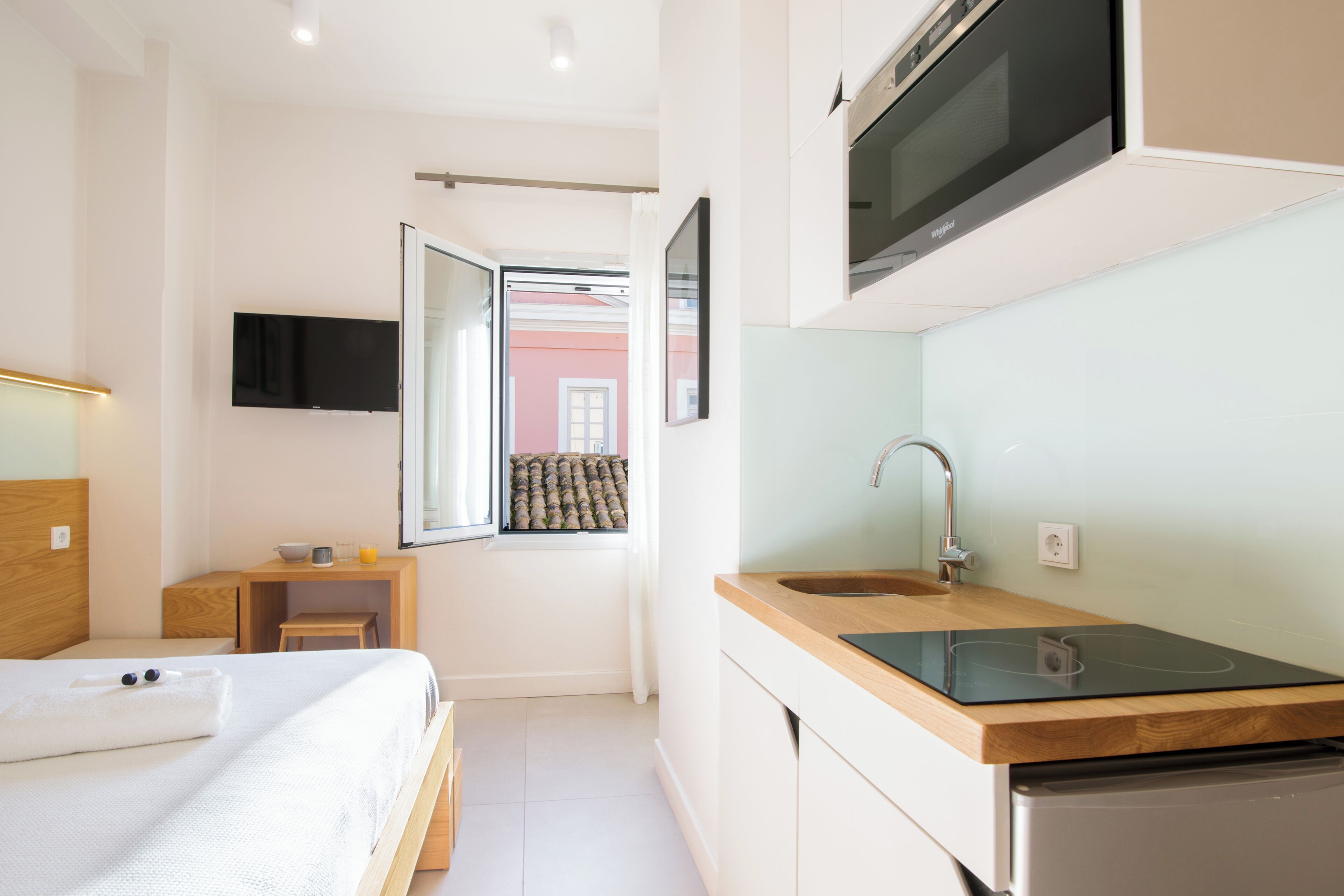 Quarto de hotel moderno com cama, pequena mesa com tigela e copos, janela com vista, cozinha com pia e fogão, e televisão de tela plana presa na parede.
