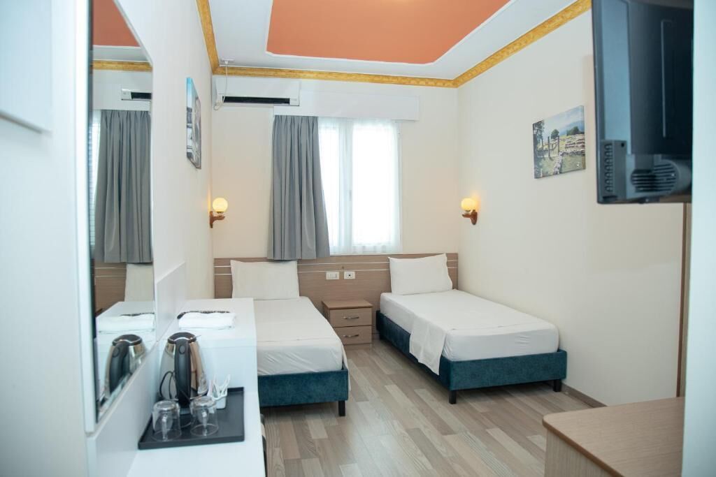Quarto de hotel com duas camas de solteiro, uma escrivaninha com cadeira, um espelho, uma televisão e uma janela com cortinas.