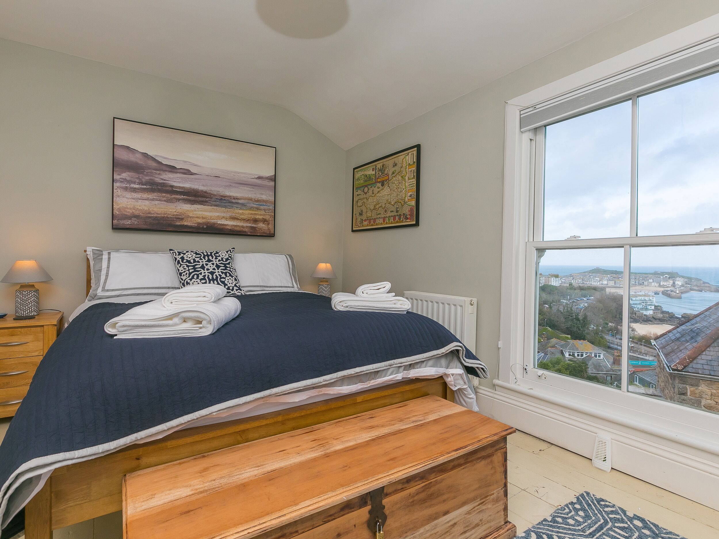 Cama bem arrumada com colcha azul escura e almofadas brancas, um banco de madeira, uma mesinha de cabeceira com abajur, e uma janela grande com vista para uma cidade costeira.