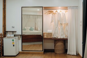 Quarto com cama, guarda-roupa cheio de roupas, pequeno frigorífico e cesto de vime.