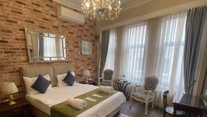 Quarto de hotel com parede de tijolos, um candelabro, cama com colcha verde, piso de madeira, mesa de jantar e uma cadeira.