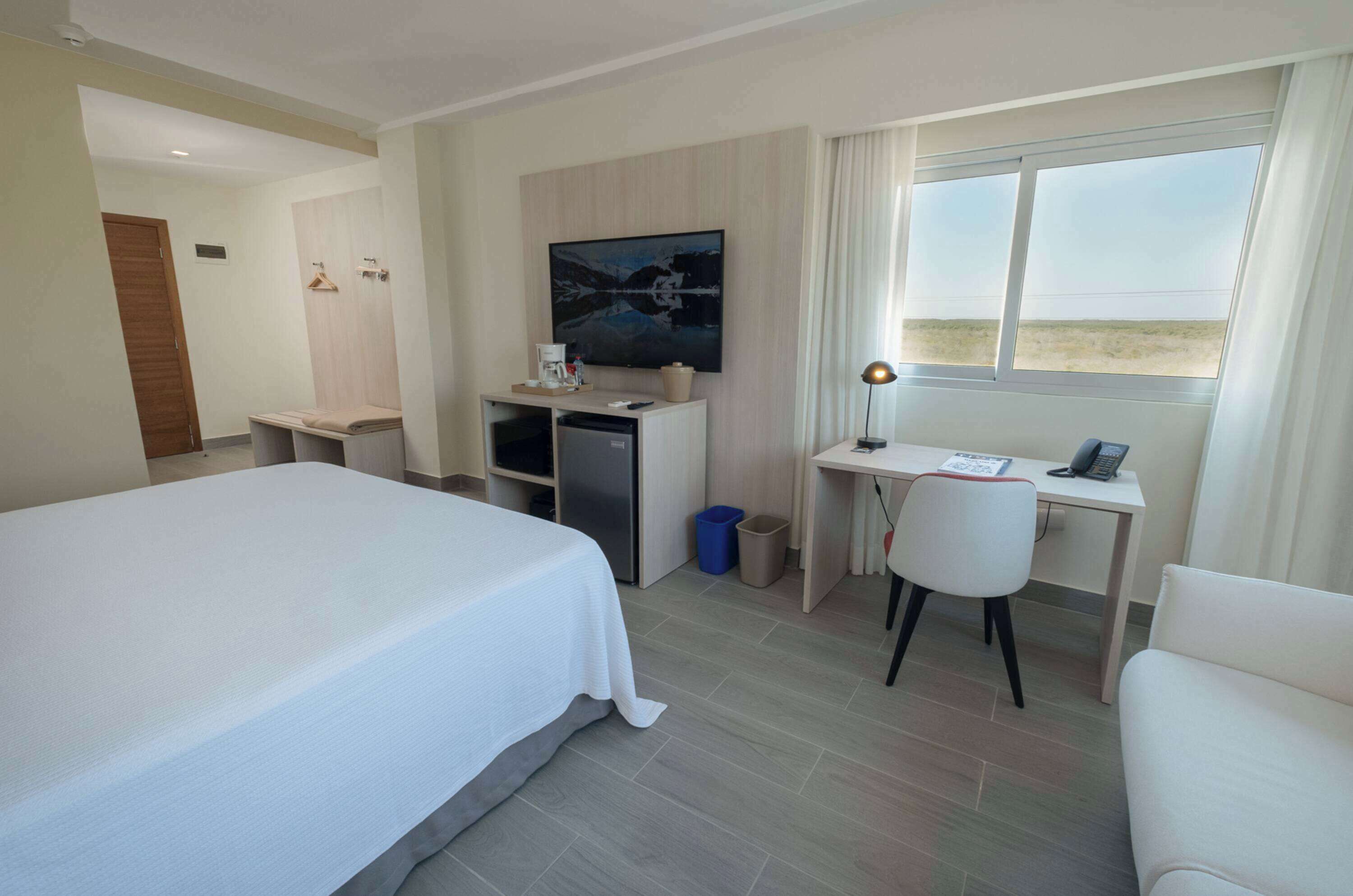 Quarto de hotel com cama, escrivaninha, cadeira e uma grande janela com vista.