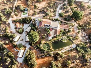 Vista aérea de uma propriedade com vários edifícios, uma construção central com telhado inclinado e um pequeno lago.
