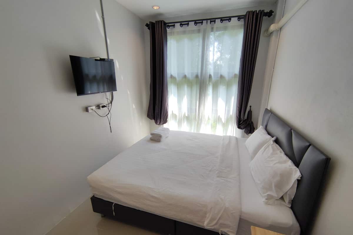 Cama bem arrumada com lençóis brancos, televisão de tela plana na parede e janela com cortinas leves.