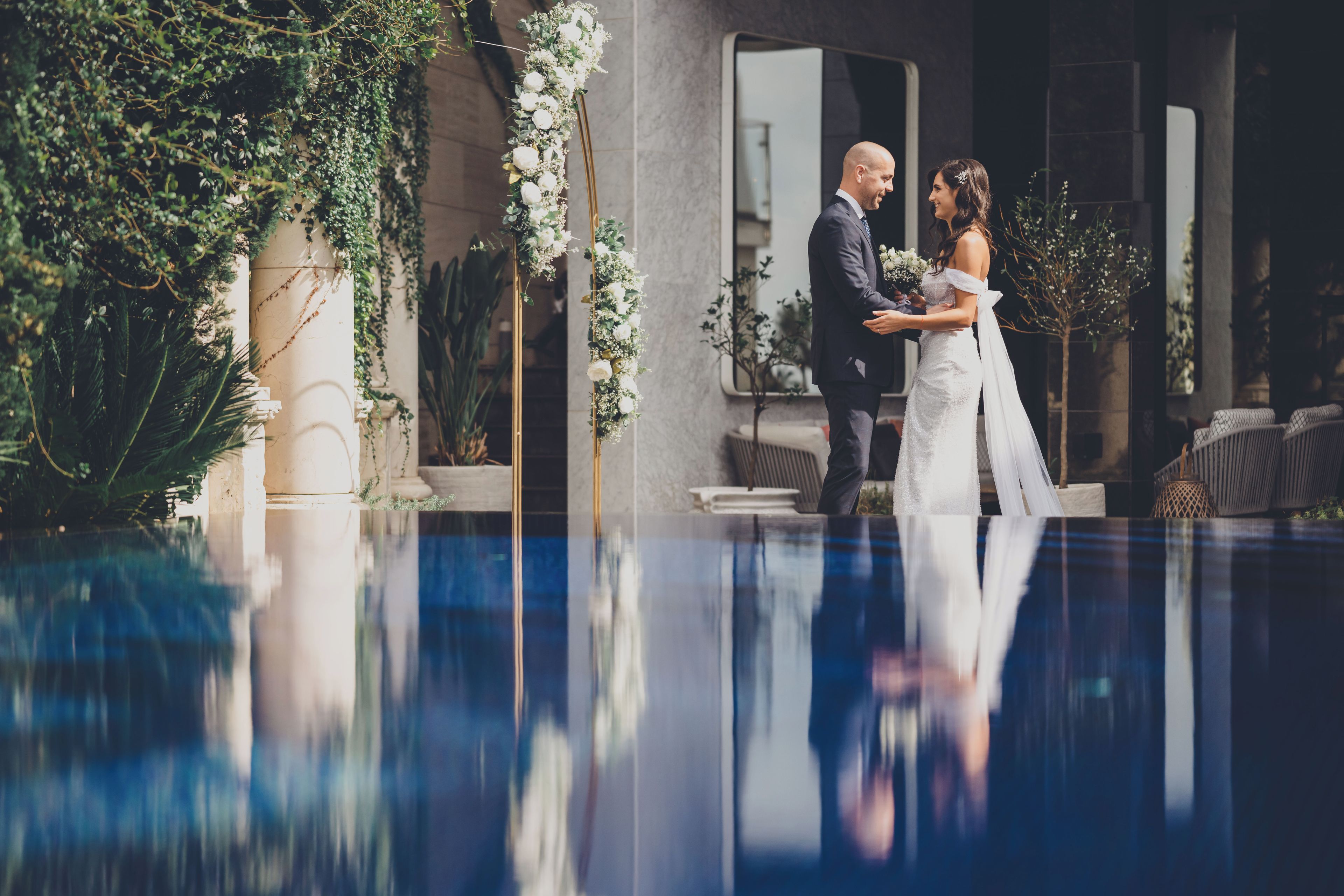Um casal está ao lado de uma piscina, com um arranjo de flores e um edifício ao fundo.