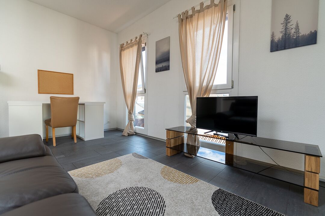 Uma sala de estar moderna com um sofá, uma televisão em um suporte de vidro, uma poltrona e uma janela com cortinas.