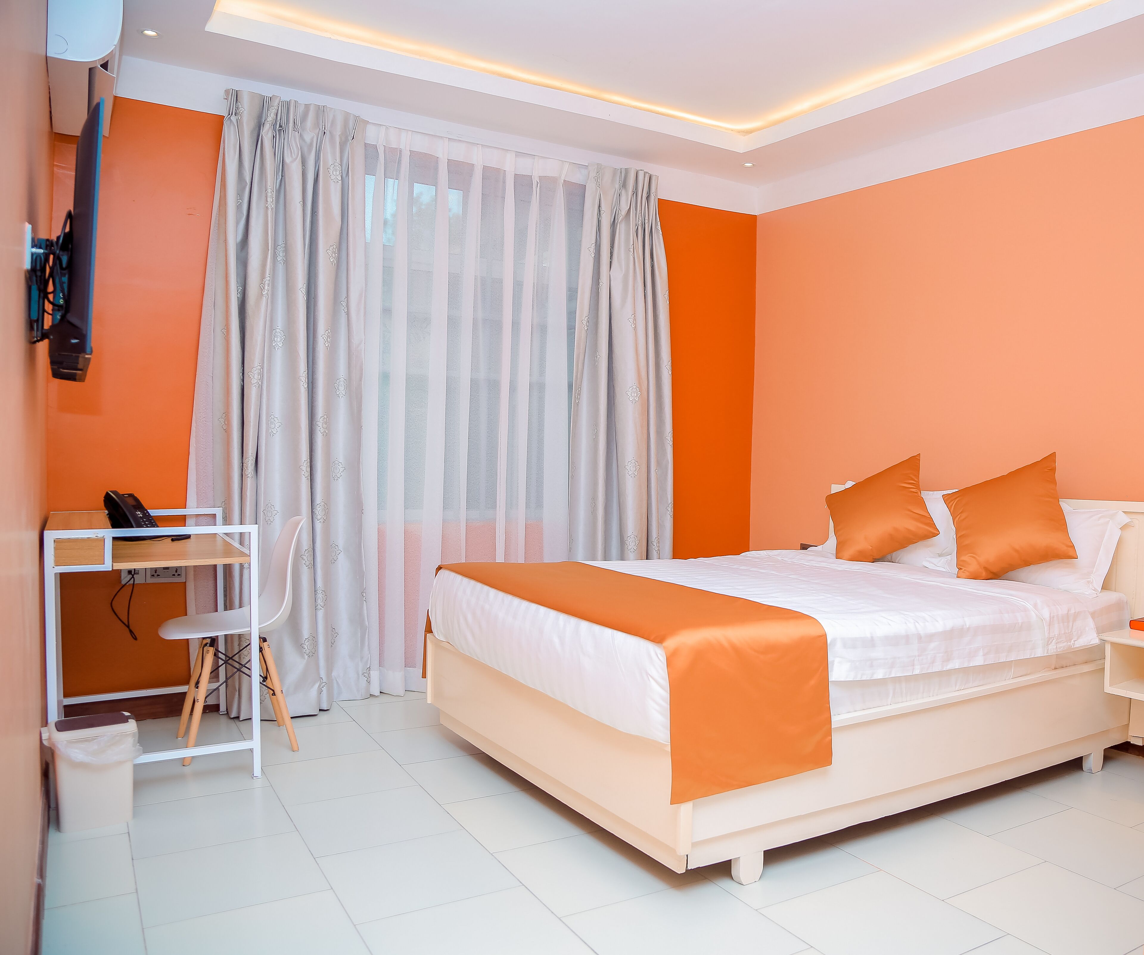 Quarto de hotel com cama, escrivaninha com telefone e janela com cortinas.