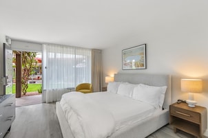 Quarto de hotel com cama, mesa de cabeceira com luminária, uma cadeira, porta que dá para uma varanda e um quadro na parede.
