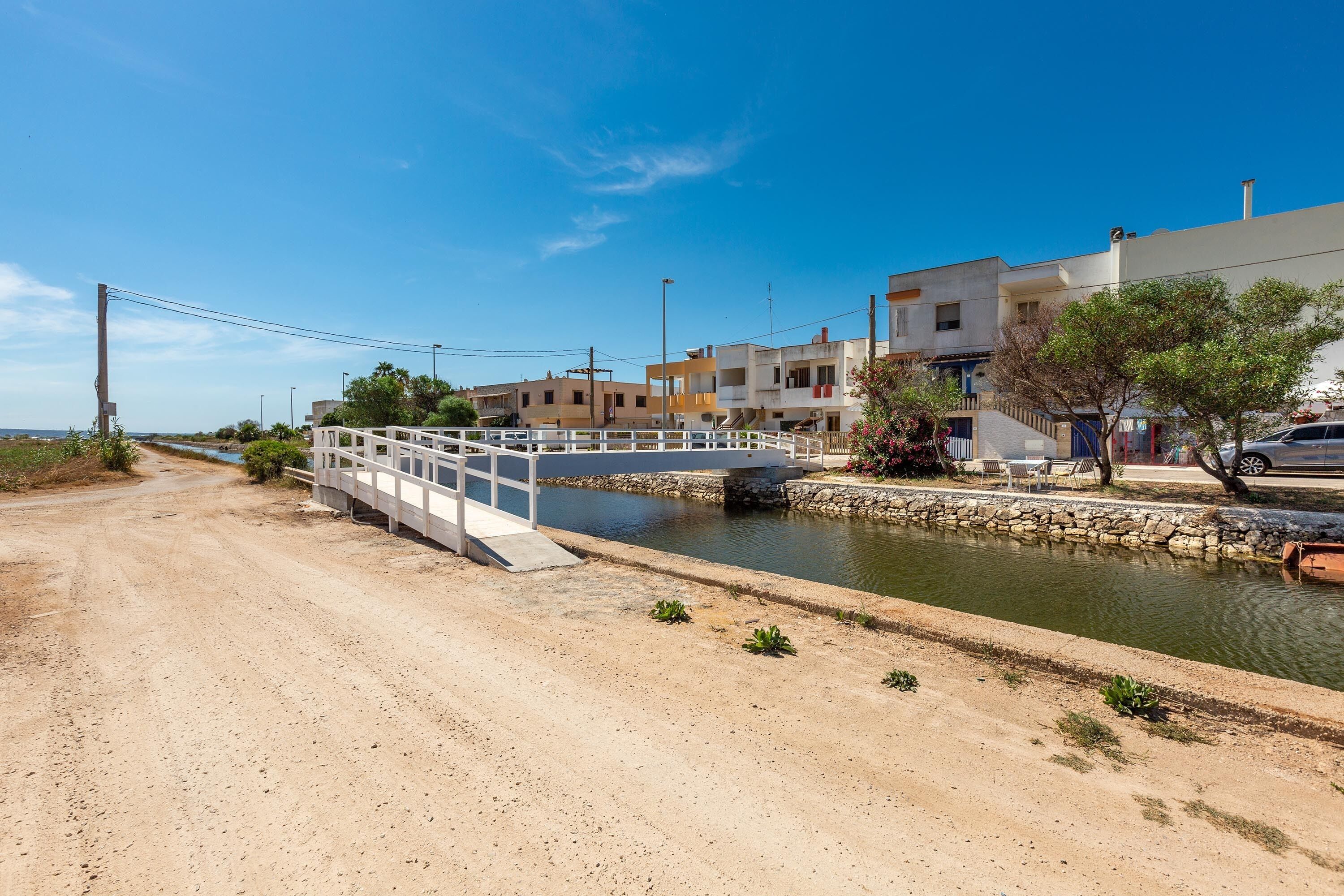 Uma estrada costeira com uma ponte branca sobre um canal, levando a uma fileira de edifícios com sacadas e um céu azul límpido.
