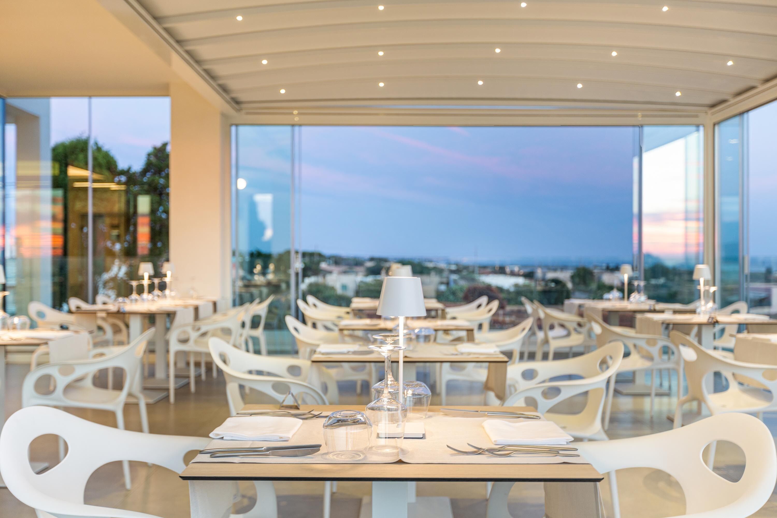 Um restaurante com cadeiras e mesas brancas, uma divisória de vidro e vista para a paisagem urbana.