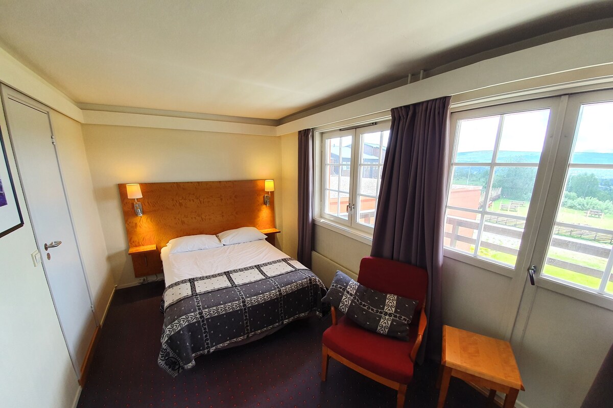 Quarto de hotel com cama, uma cadeira, mesa pequena, janela com cortinas e luminária na parede.