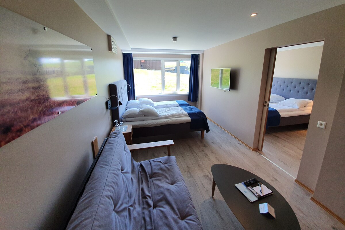 Quarto de hotel moderno com uma cama, um sofá, uma mesinha, uma televisão e uma porta que dá para outro quarto.