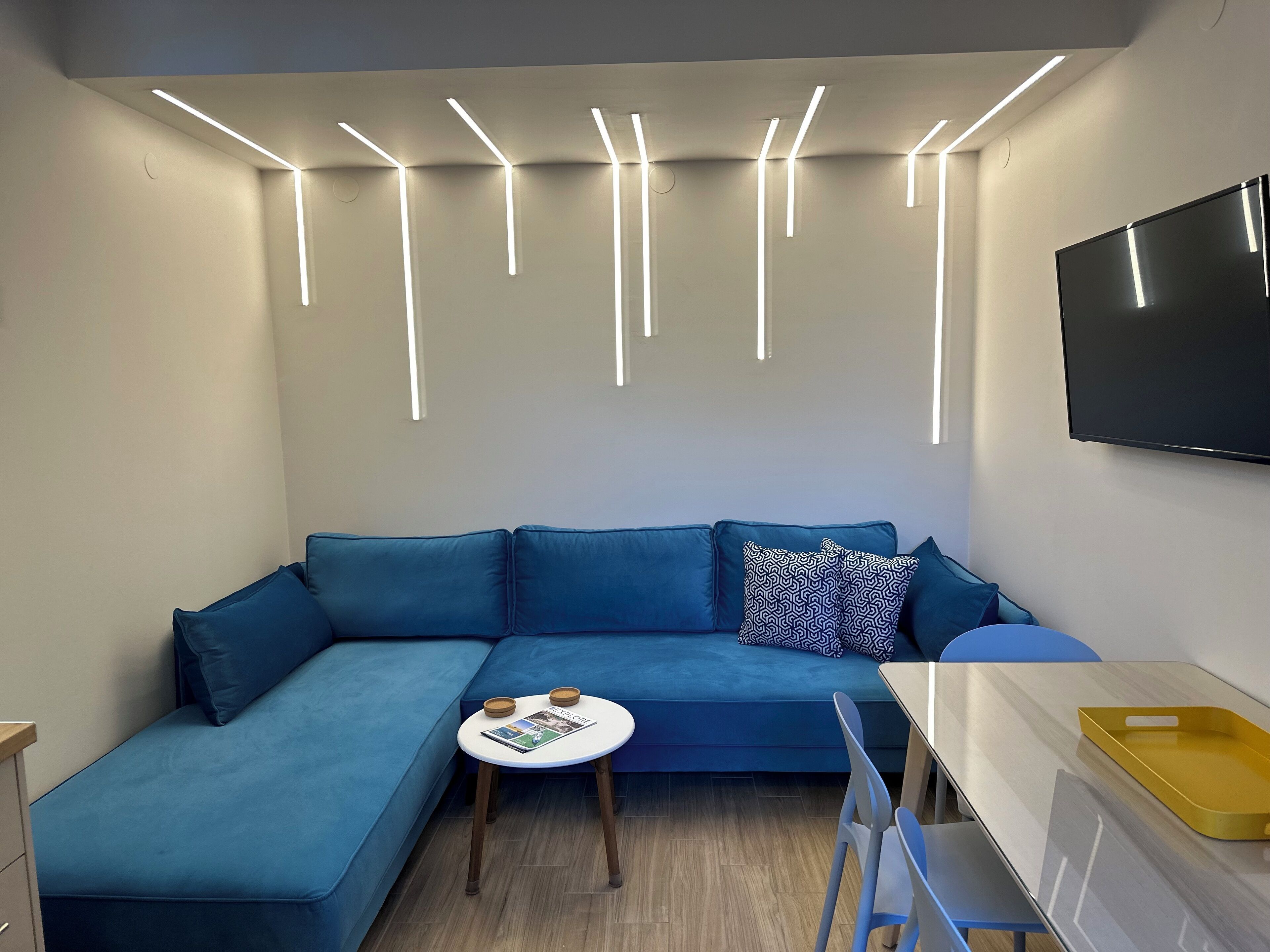 Uma sala de estar moderna com um sofá seccional azul, uma mesa redonda pequena com revistas, uma TV de tela plana na parede e uma bandeja amarela sobre um armário branco.