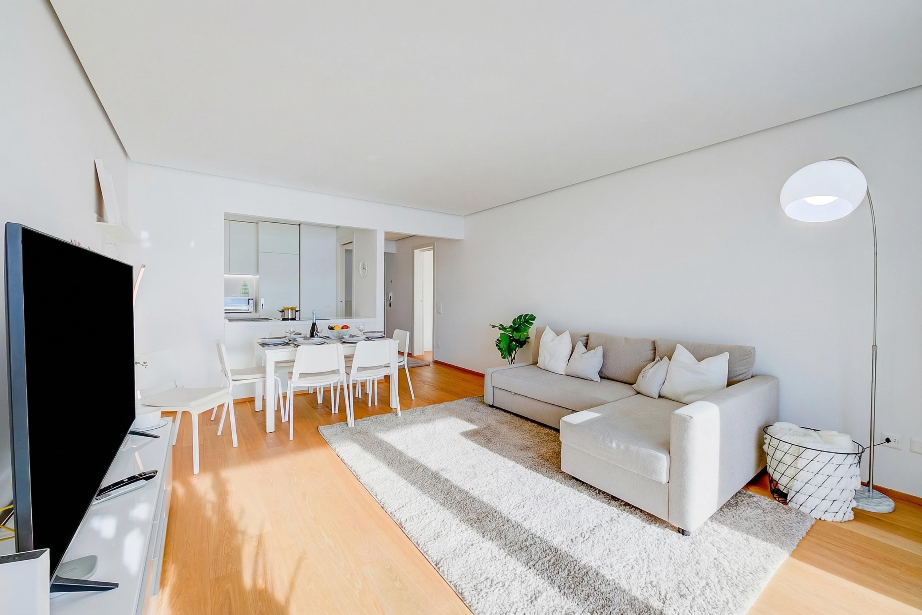 Uma sala de estar moderna com sofá, televisão, área de jantar com cadeiras e um abajur.