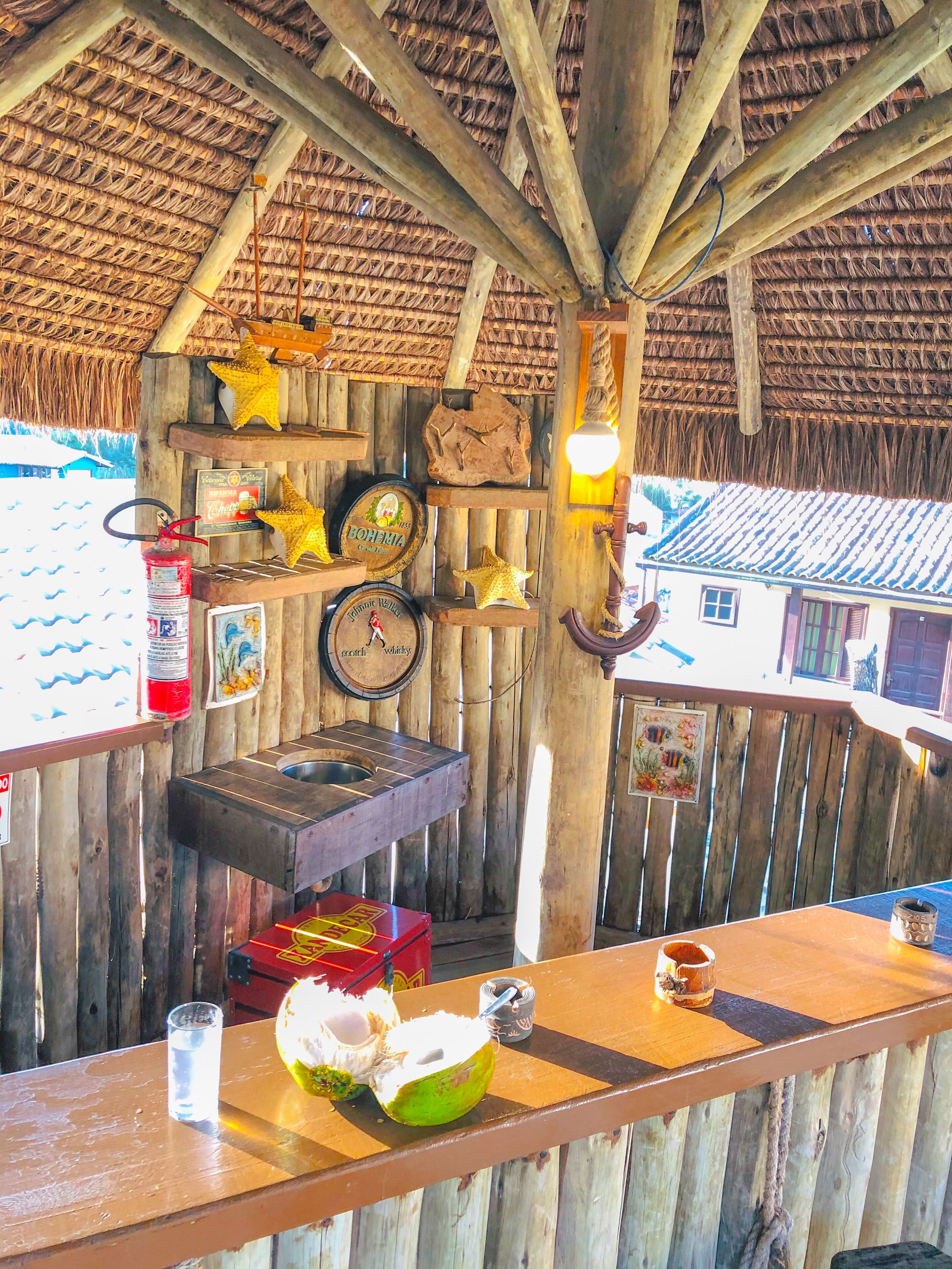 Um espaço de bar rústico com paredes de madeira, um telhado de palha e diversas decorações, incluindo estrelas-do-mar e um relógio.