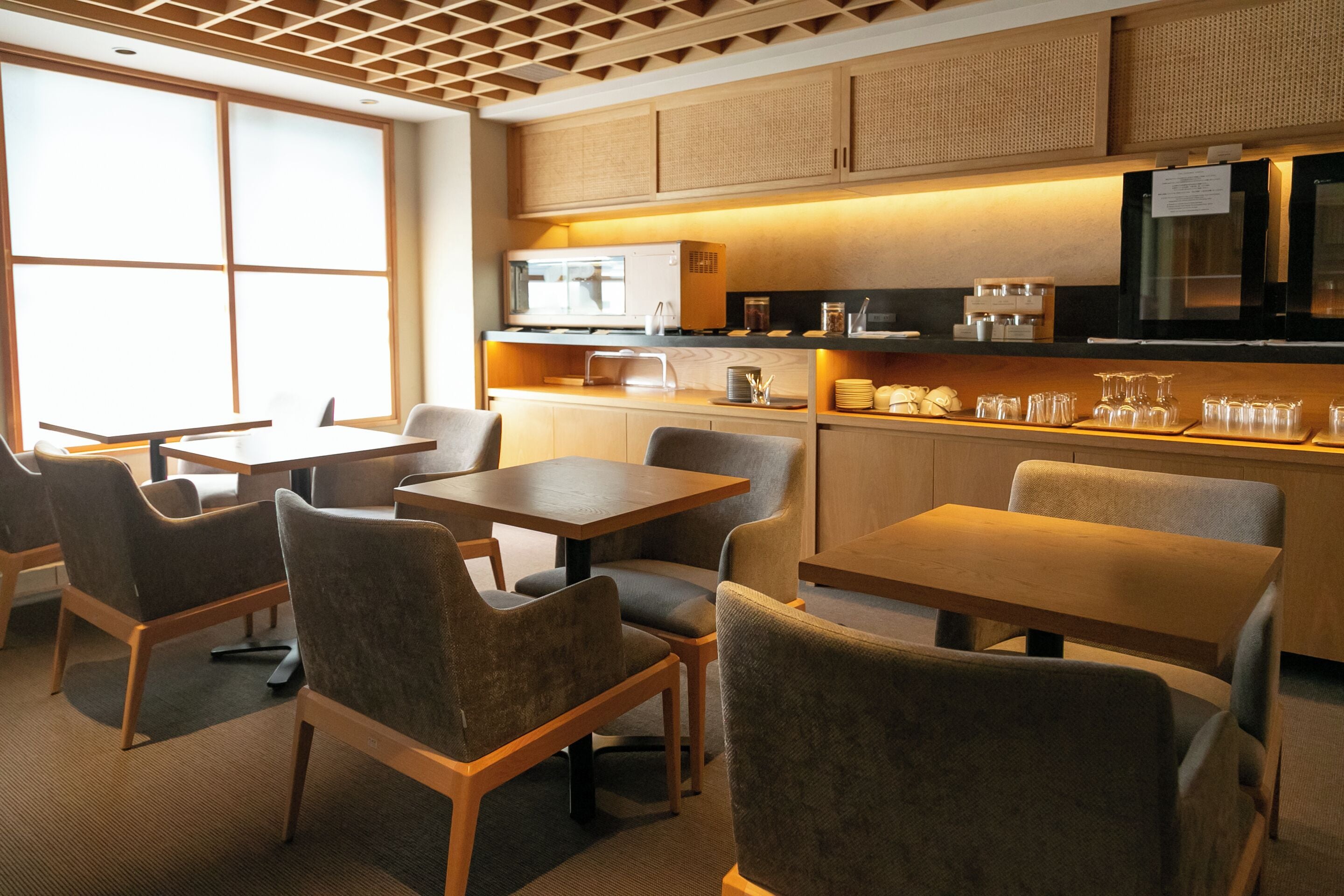 Uma sala de jantar moderna com mesas e cadeiras de madeira, área de cozinha com eletrodomésticos e grandes janelas que proporcionam luz natural.