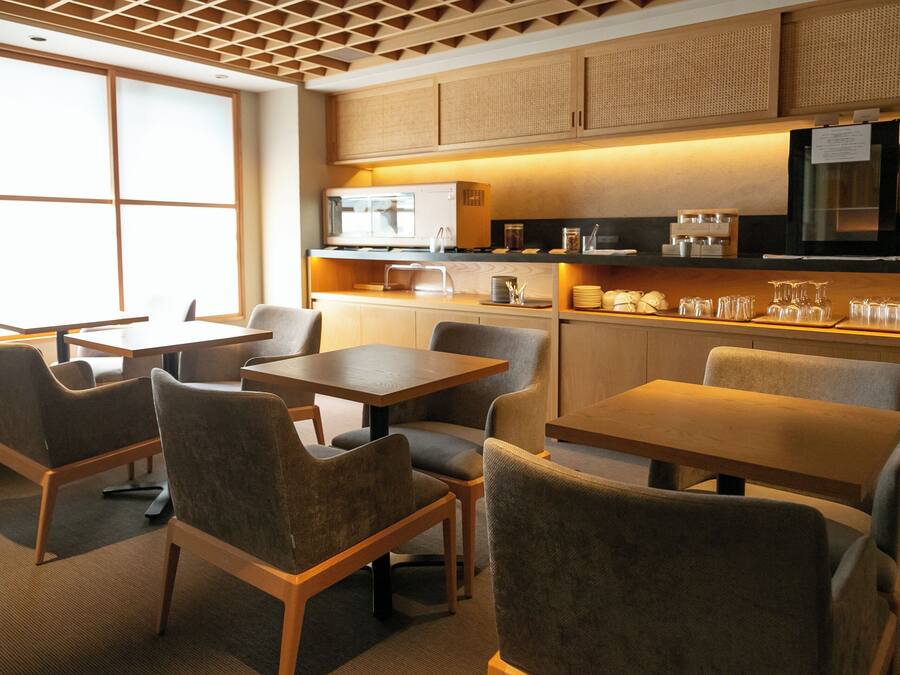 Uma sala de jantar moderna com mesas e cadeiras de madeira, área de cozinha com eletrodomésticos e grandes janelas que proporcionam luz natural.