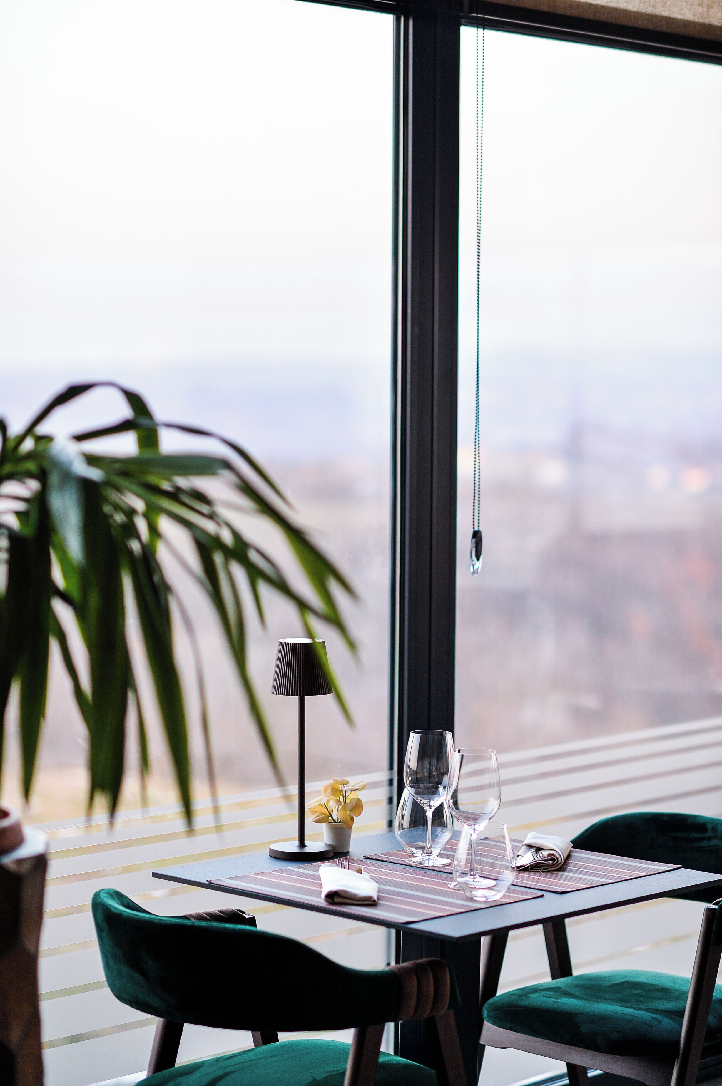 Uma sala de jantar com uma mesa posta para dois, um vaso de planta e vista da cidade através de grandes janelas.