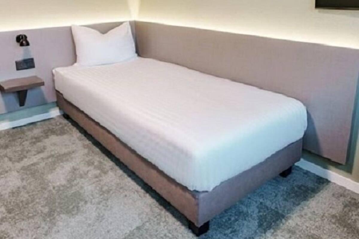 Uma cama de solteiro com cabeceira, mesa de cabeceira e luminária fixa na parede.