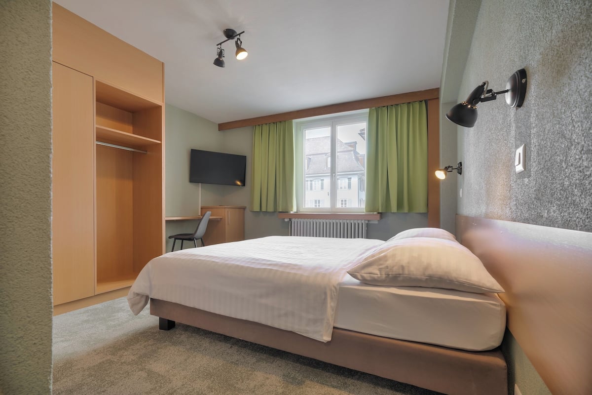 Quarto com cama, escrivaninha, cadeira, guarda-roupa, televisão e janela com cortinas.