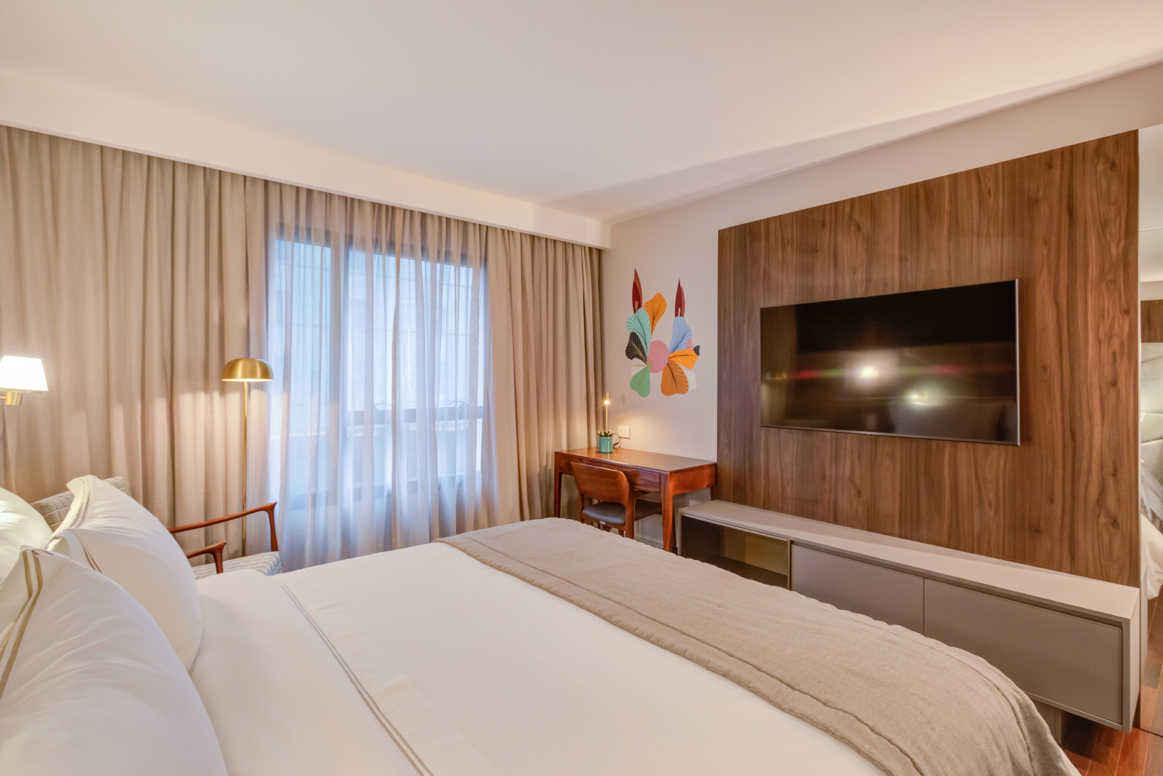 Quarto de hotel com uma cama grande, uma escrivaninha, televisão na parede e janela com cortinas.