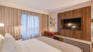 Quarto de hotel com uma cama grande, uma escrivaninha, televisão na parede e janela com cortinas.