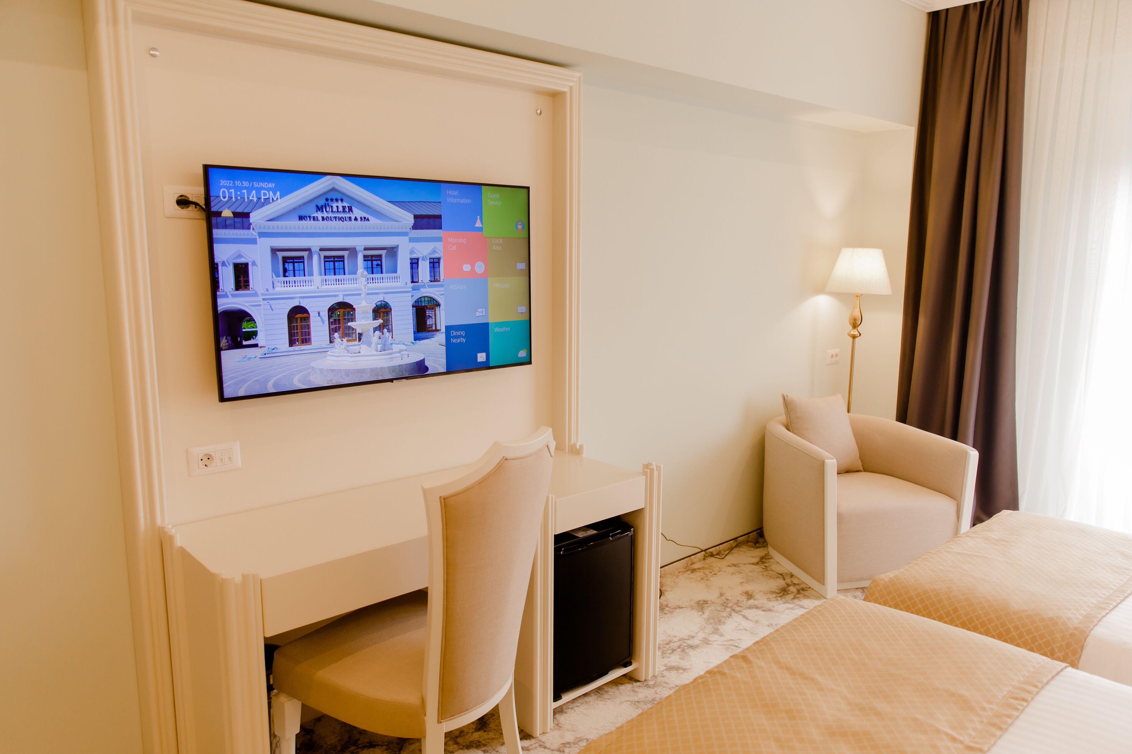 Quarto de hotel com televisão na parede exibindo uma imagem do hotel, uma mesa com cadeira, um sofá e um abajur.