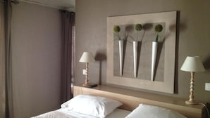 Quarto de hotel com cama, duas luminárias de cabeceira, uma obra de arte na parede e uma janela com cortinas.