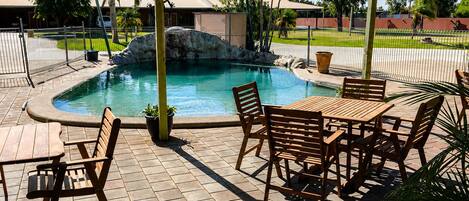 Uma área de piscina com mesa e cadeiras de madeira, cercada por uma grade e palmeiras.
