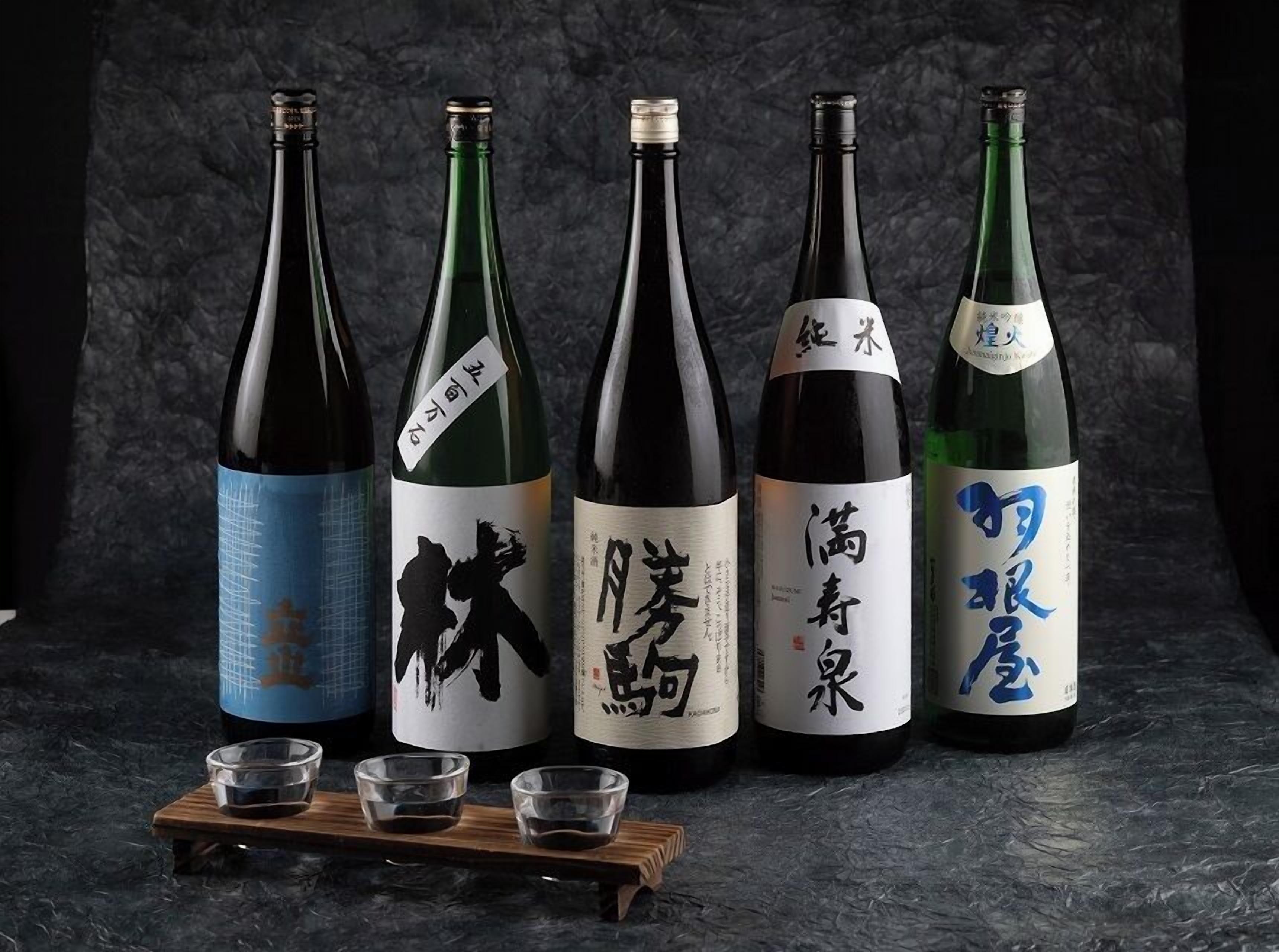 Cinco garrafas de saquê com etiquetas japonesas, cada uma acompanhada por uma pequena taça.