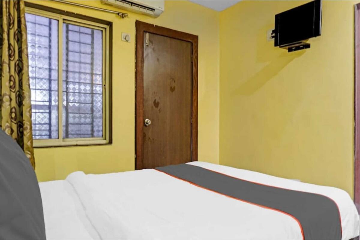 Quarto com cama, janela com cortinas, porta e televisão montada na parede.