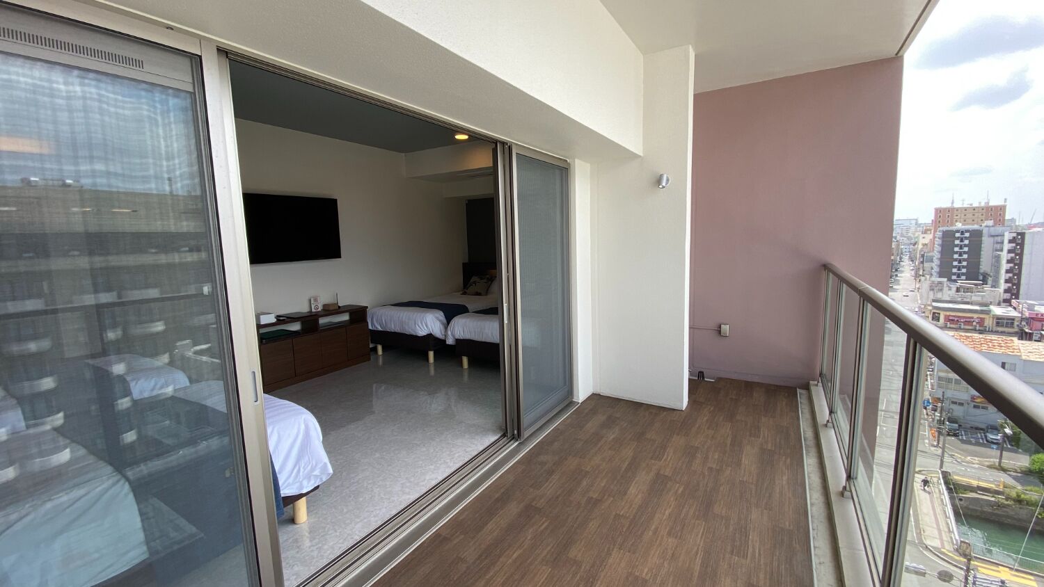 Varanda com vista para a cidade, porta de correr de vidro que dá para um quarto com duas camas e uma televisão fixada na parede.