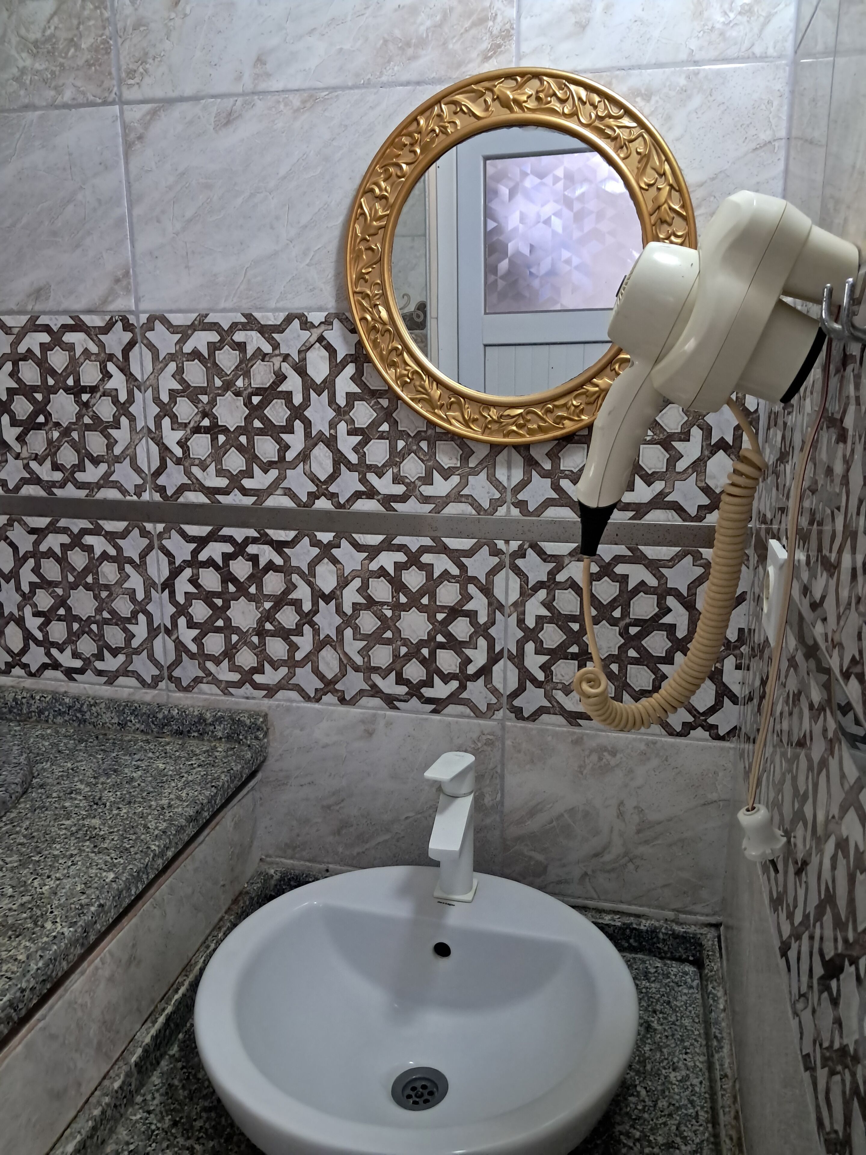 Casa de banho com lavatório, espelho com moldura decorativa e secador de cabelo fixado na parede.