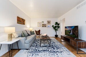 Uma sala de estar moderna com sofá, mesa de centro e TV. Há obras de arte nas paredes e um vaso de planta.