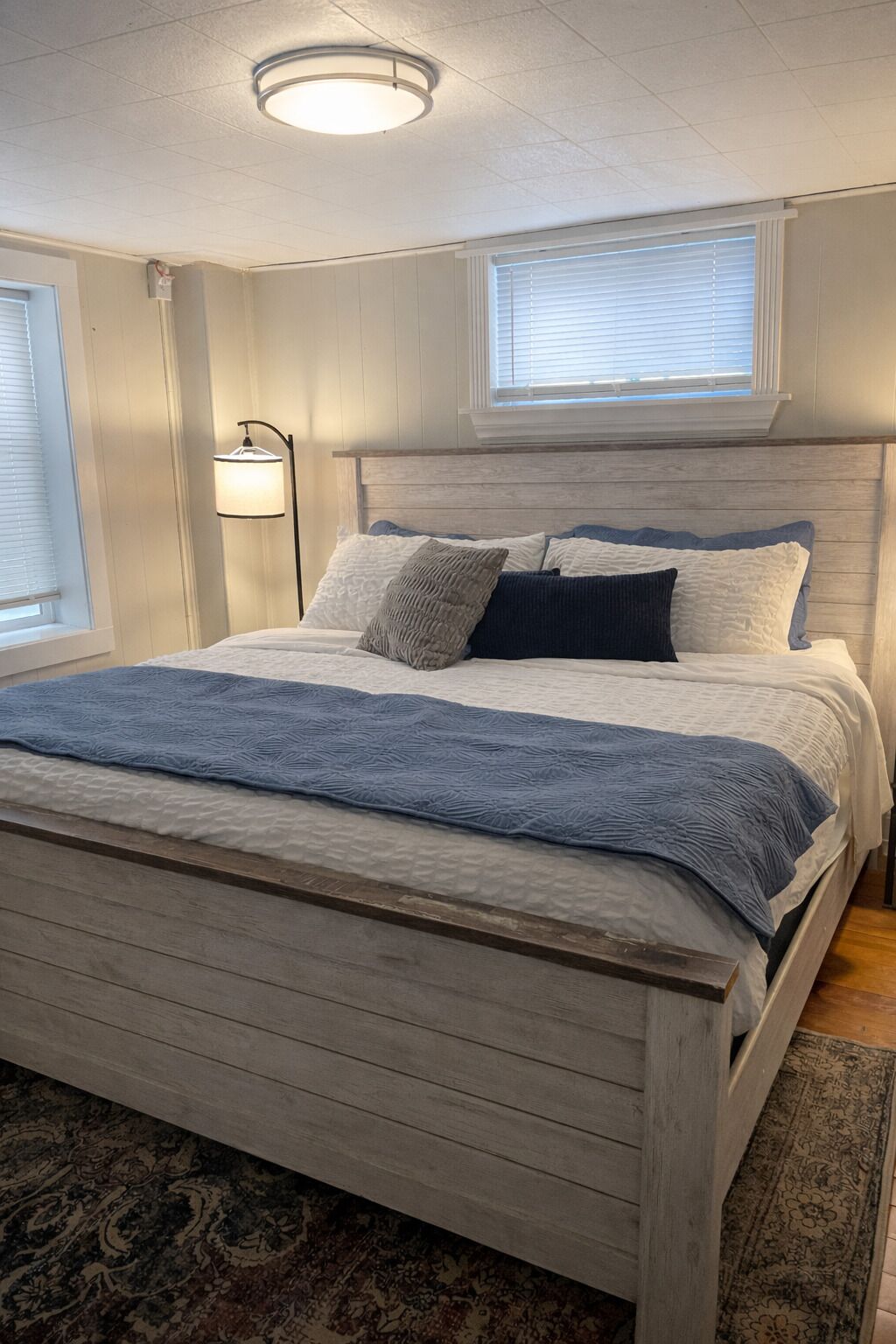Quarto com cama de madeira, mesa de cabeceira com abajur, janela com persianas e tapete estampado.