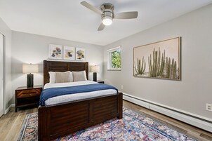 Um quarto com uma cama de madeira, duas mesas de cabeceira, um ventilador de teto e uma grande pintura de cacto na parede.