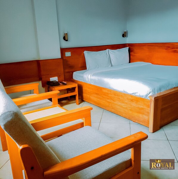 Quarto de hotel com uma cama e uma cadeira de madeira, mesa de cabeceira com telefone e tomada na parede.