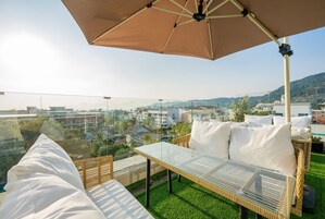 Uma varanda com mesa de vidro, sofá de vime e um grande guarda-sol.